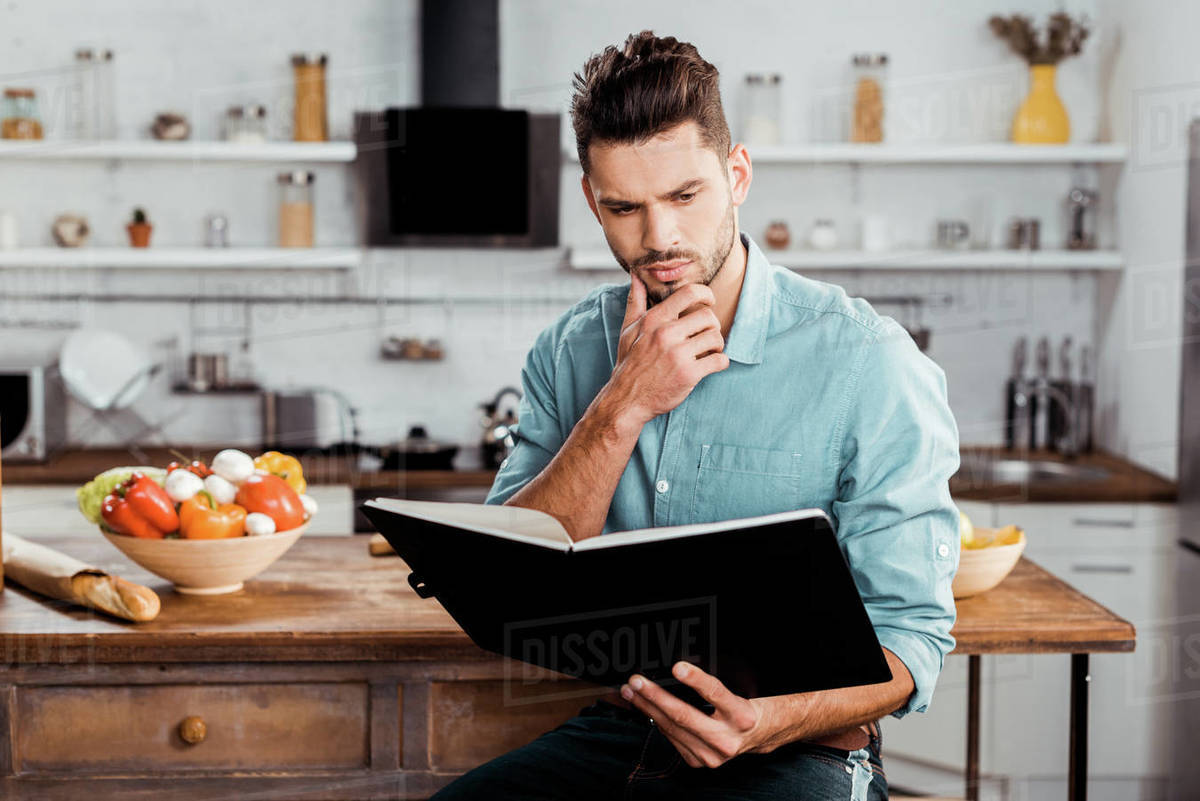Thoughtful handsome young man reading cookbook in kitchen - Royalty ...