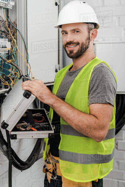 Smiling handsome electrician holding toolbox near electrical box in ...