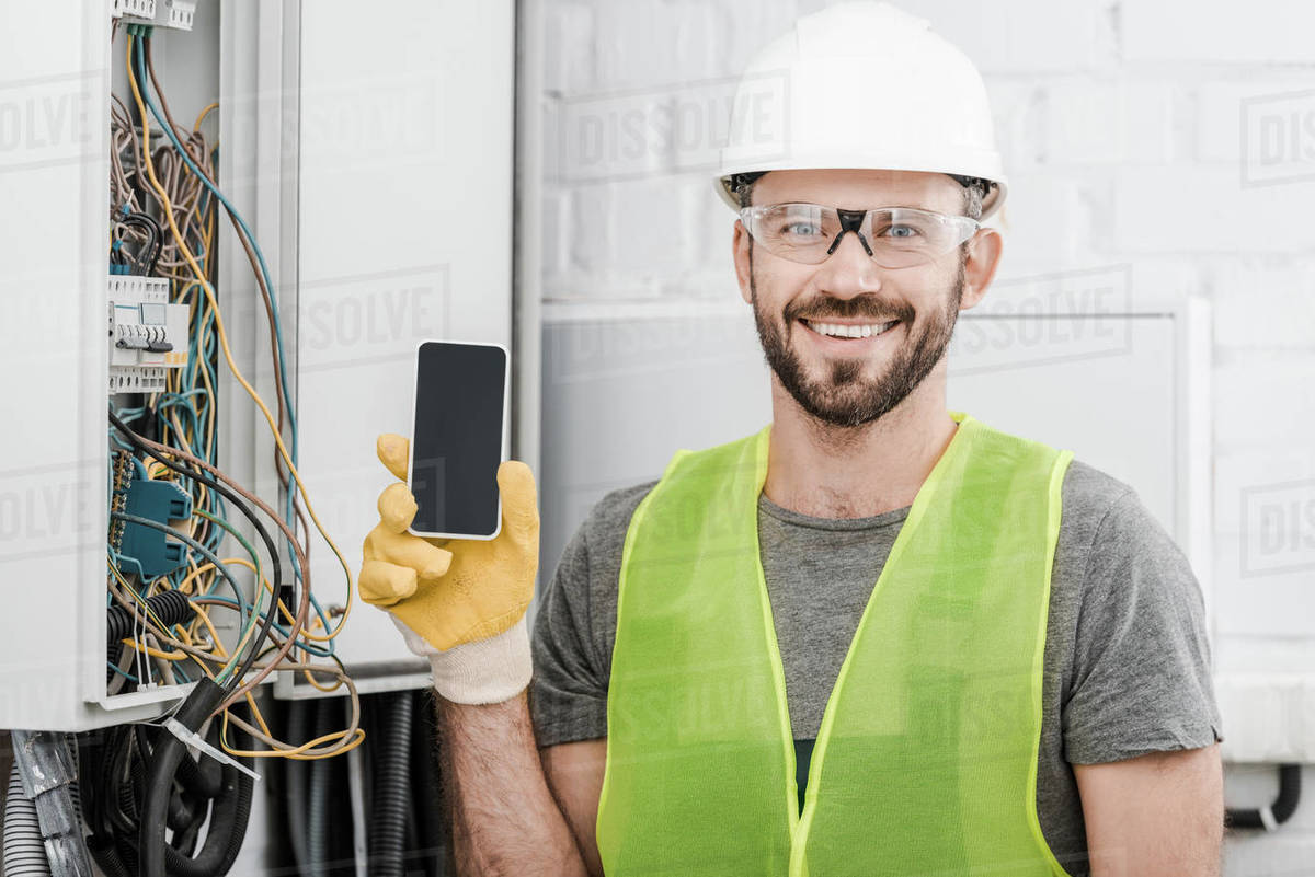 Smiling handsome electrician showing smartphone with blank screen near ...