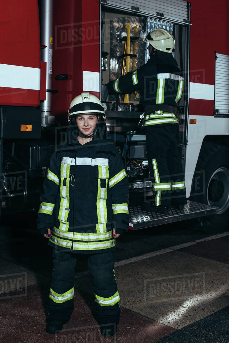 smiling female firefighter looking at camera while colleague checking ...