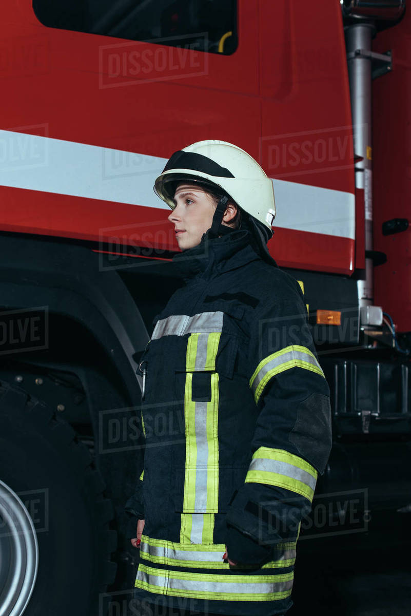 side view of female firefighter in helmet standing at fire station ...