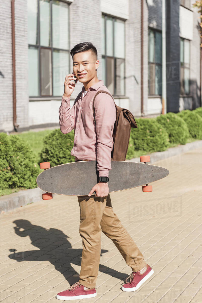 Side view of smiling Asian man with longboard having conversation on ...