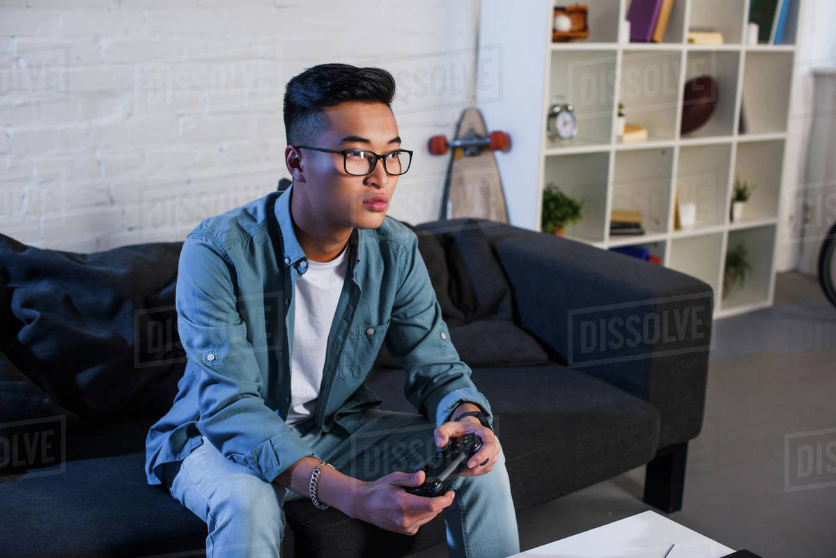 Focused young Asian man playing video game with joystick at home ...