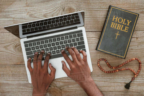 Cropped shot of man working with laptop with holy bible and beads on ...