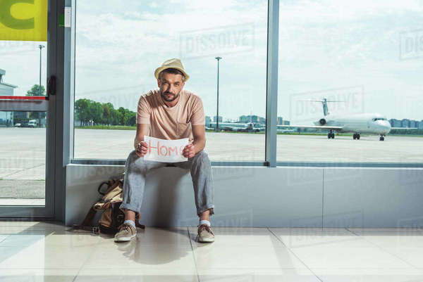 Sad tourist holding card with home word while sitting in airport ...