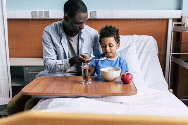 Portrait of African American father feeding little sick son in clinic ...