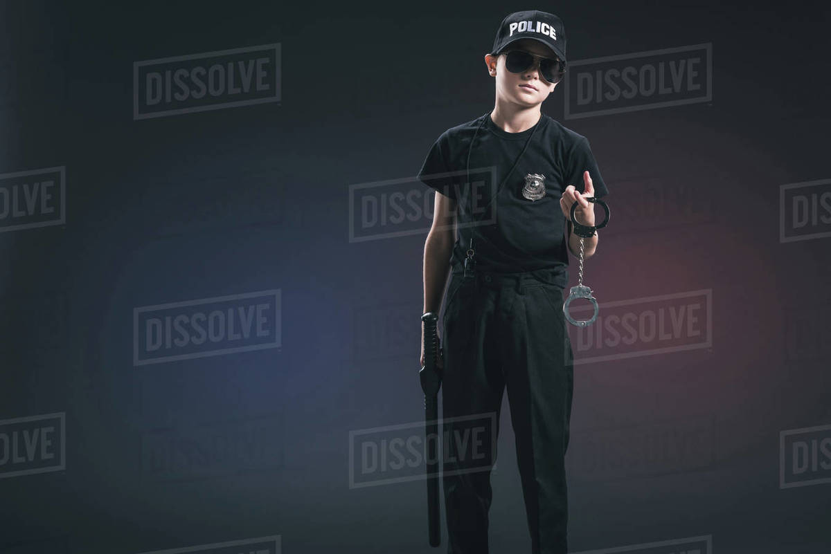 Boy in policeman uniform and sunglasses with handcuffs on dark ...