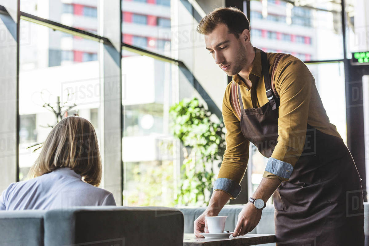 Confident young waiter serving cup of coffee for female client at cafe ...
