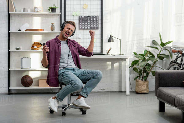 Happy man listening music and singing while sitting on chair at home ...