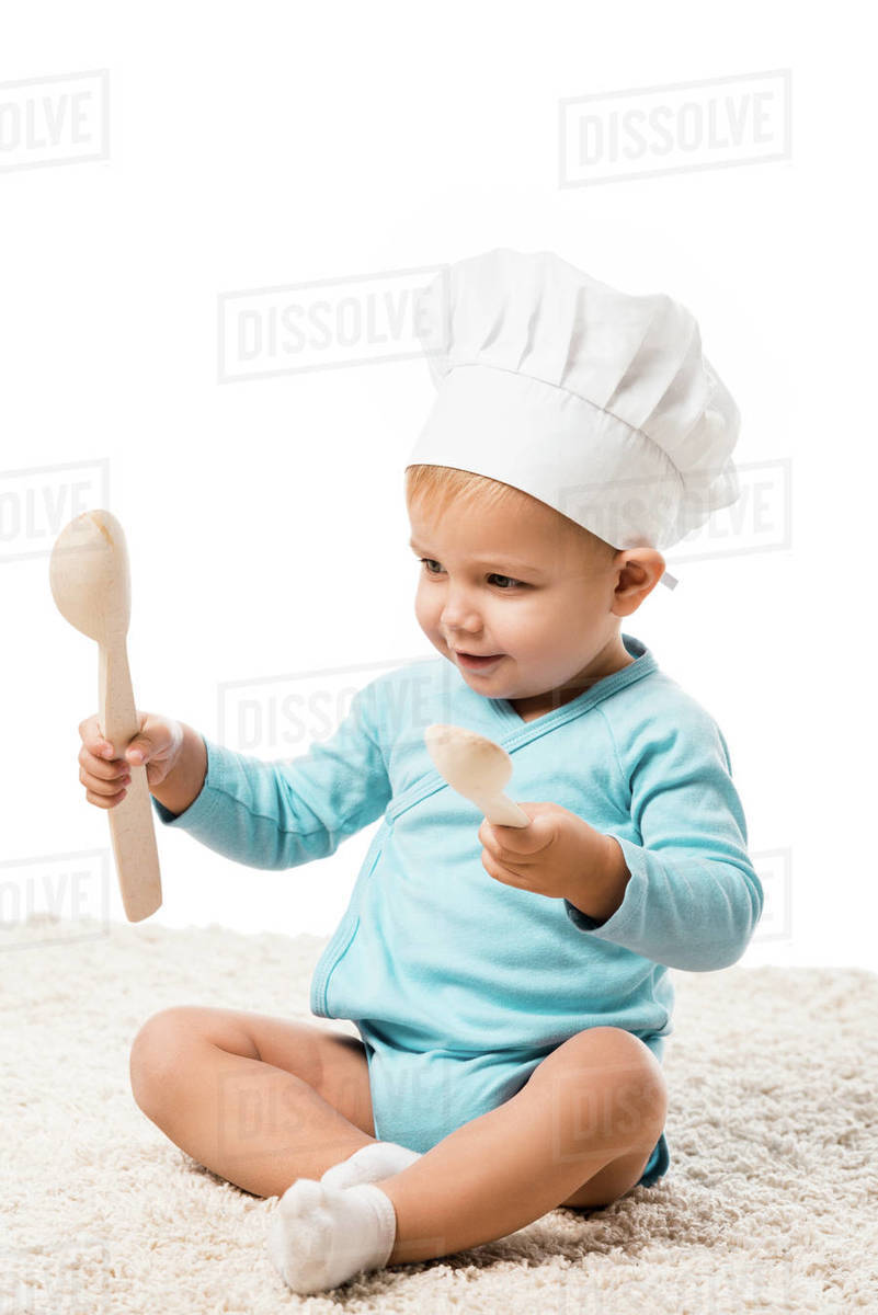 Toddler boy in chefs hat sitting on carpet, holding two big wooden ...