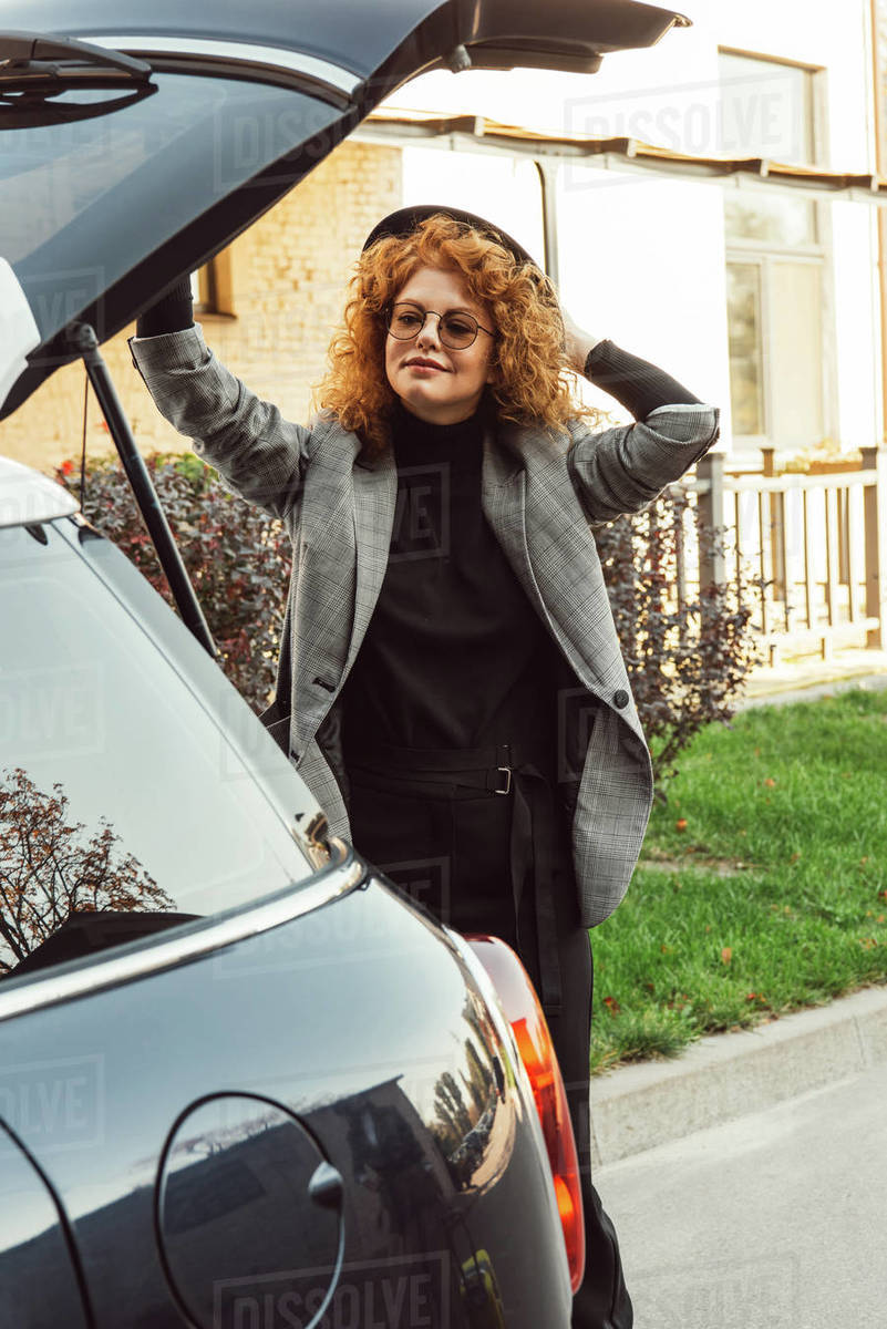 Stylish curly redhead woman in grey jacket closing car trunk at city ...