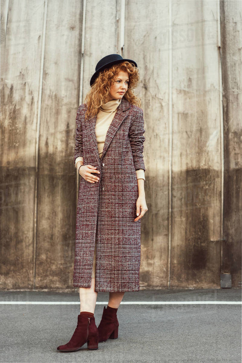 Adult curly redhead woman in black hat and coat posing at city street ...