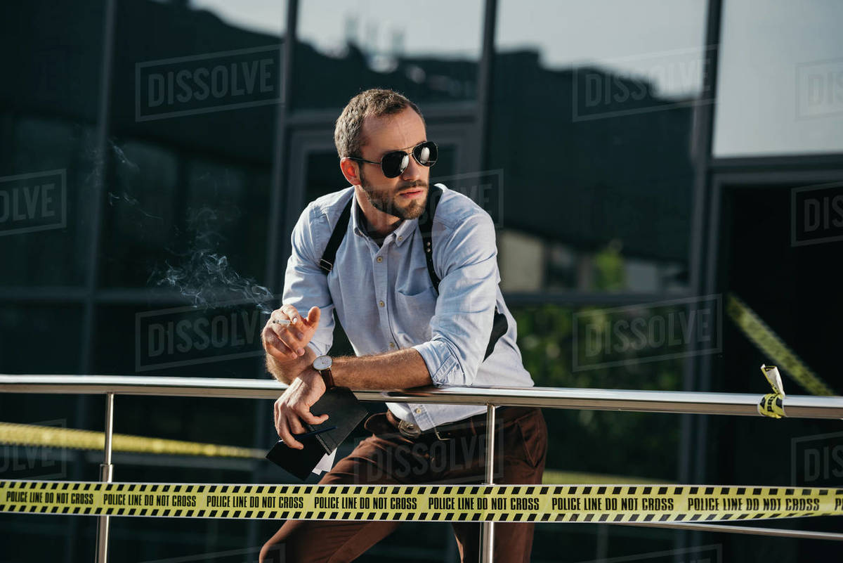 Male detective in sunglasses smoking and looking to the side leaning on ...