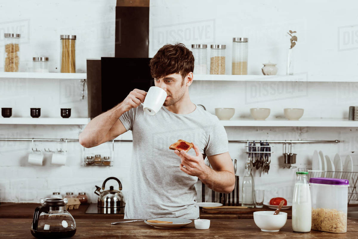 young man drinking coffee and eating toast with jam on breakfast at ...