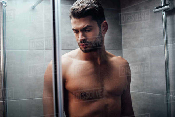 Selective focus of serious handsome man standing behind glass shower ...