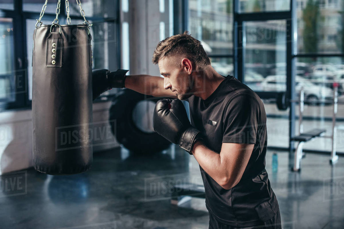 Side view of handsome muscular boxer training with punching bag in gym ...