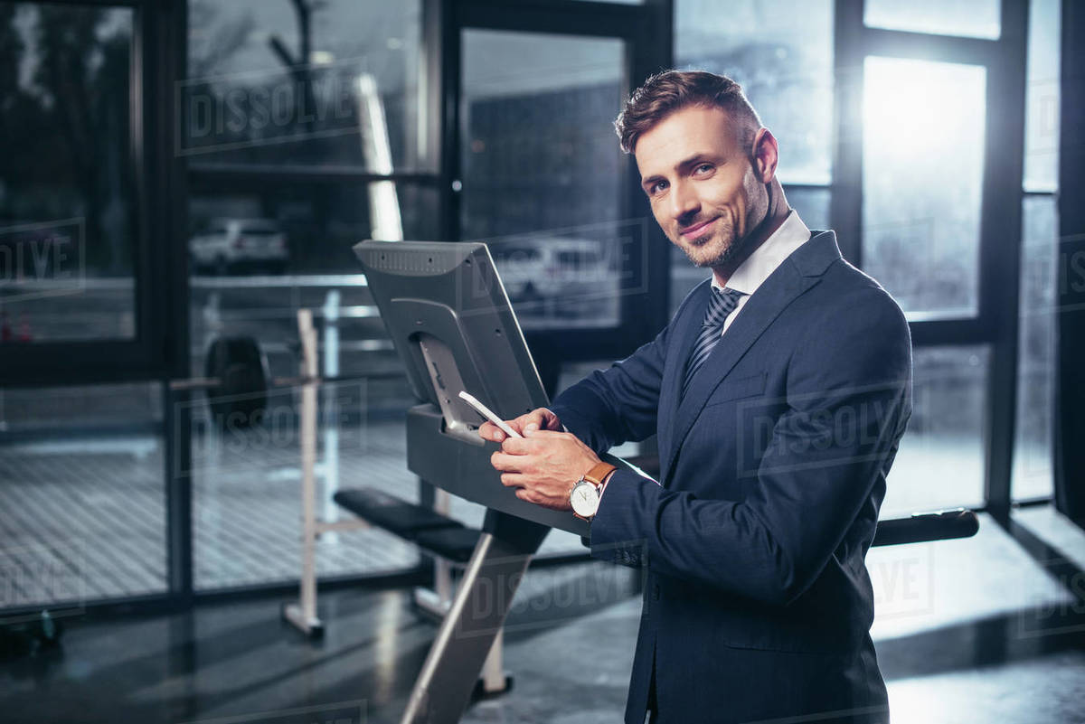 Handsome businessman in suit leaning on treadmill and using smartphone ...