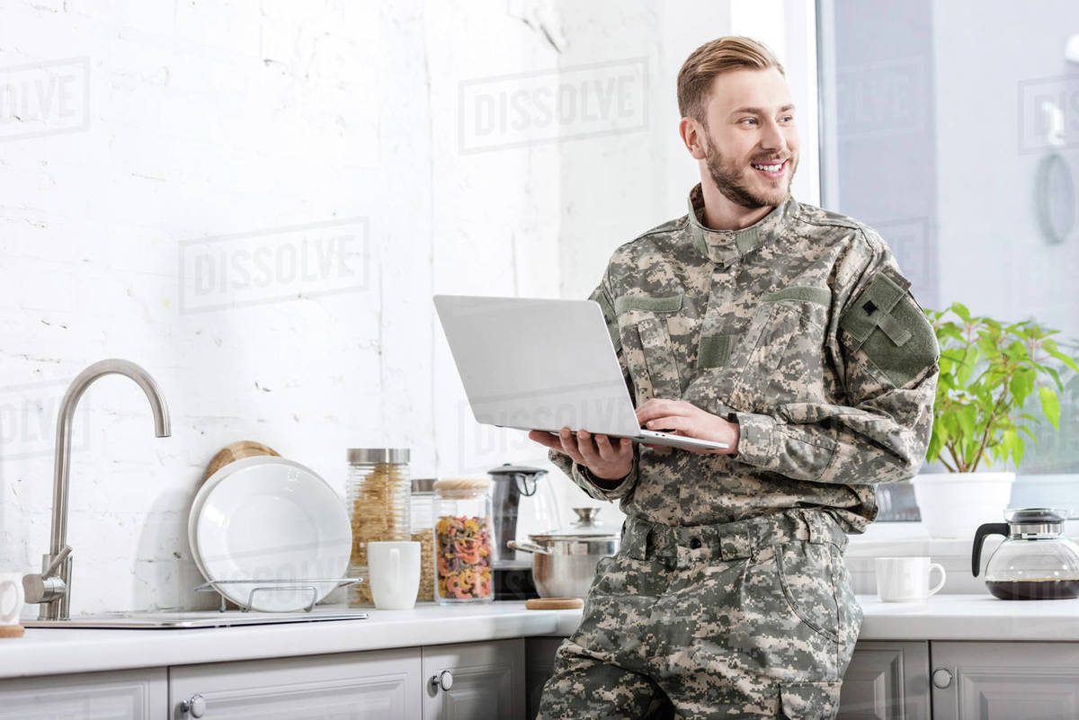 smiling army soldier using laptop at kitchen and looking away - Royalty ...