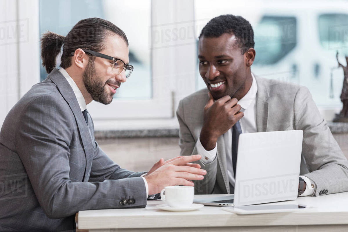 smiling multicultural businessmen sitting at table with laptop and ...