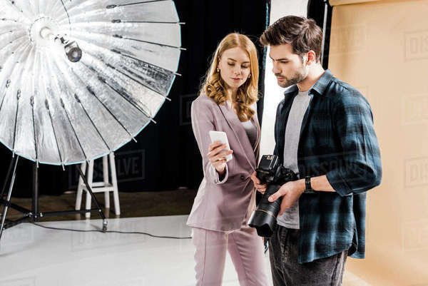 Young photographer with camera and beautiful model using smartphone in ...