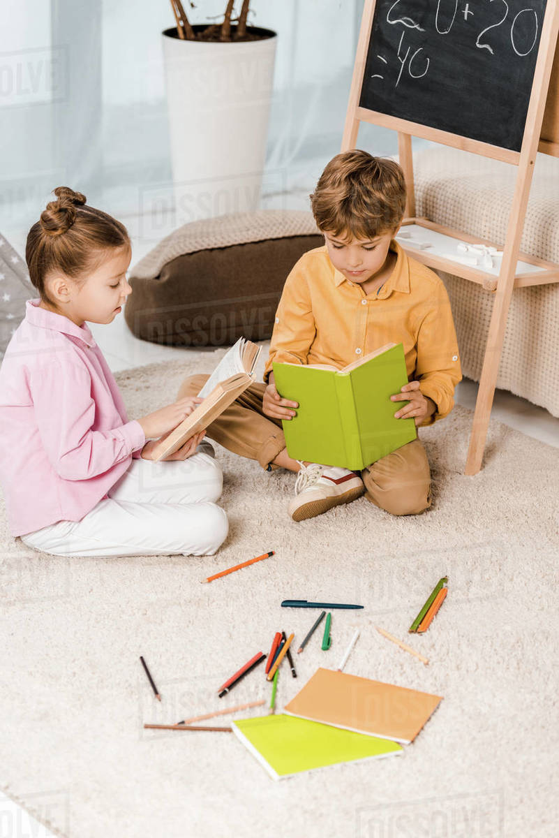 High angle view of adorable focused kids sitting on carpet and reading ...
