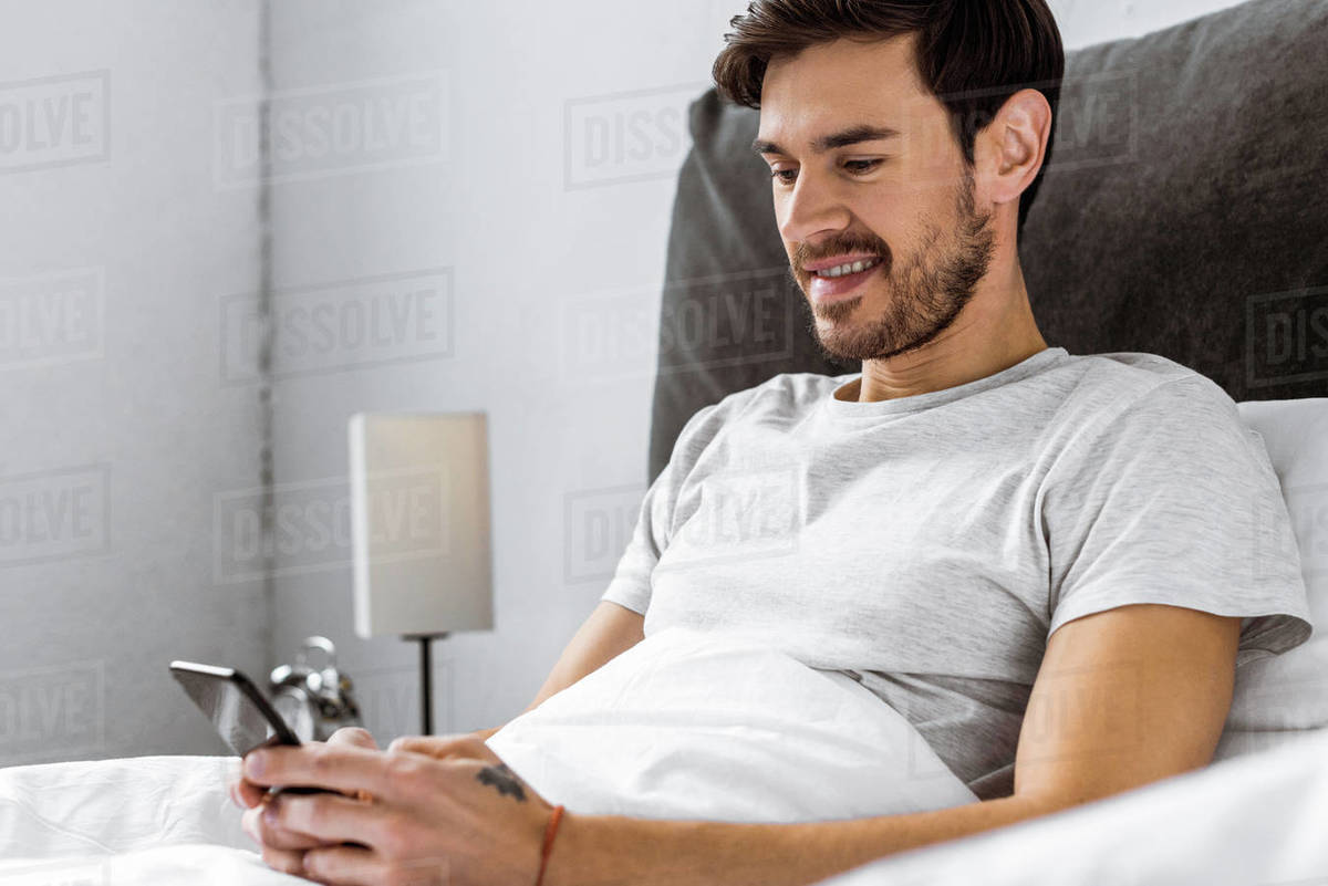 handsome smiling young man sitting in bed and using smartphone ...