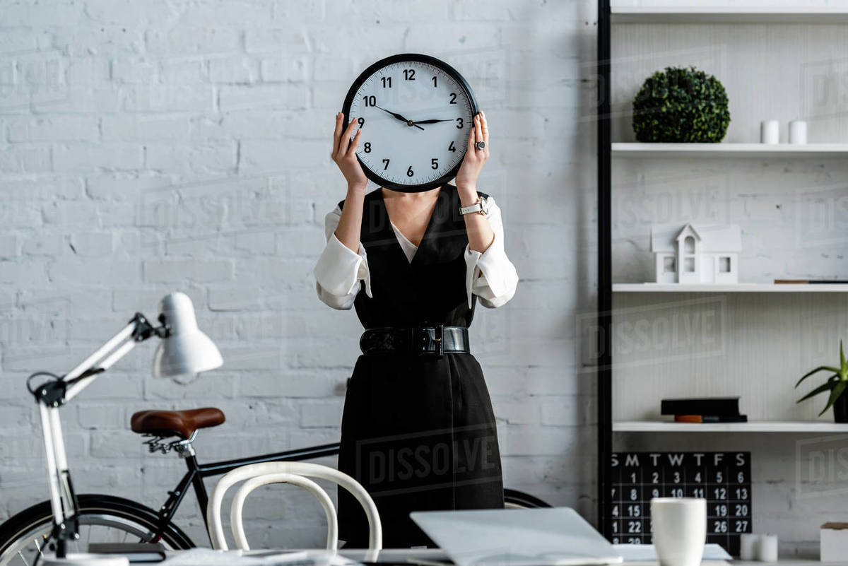 Businesswoman in formal wear holding clock in front of face in office ...