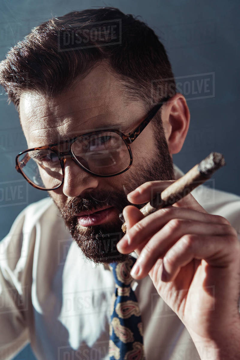 Handsome man in glasses holding cigar and looking at camera on grey ...