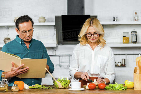 Mature wife and husband cooking together with recipe book in kitchen ...