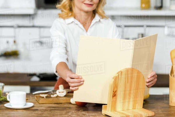 Cropped image of mature woman taking recipe book for cooking in kitchen ...