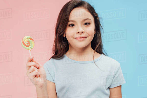 Adorable child holding candy while looking at camera on blue and pink ...