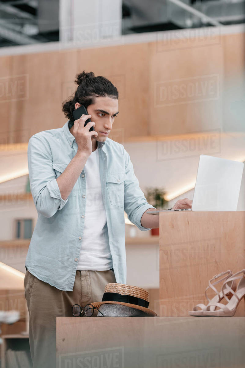 Concentrated young shop owner talking on smartphone and using laptop ...