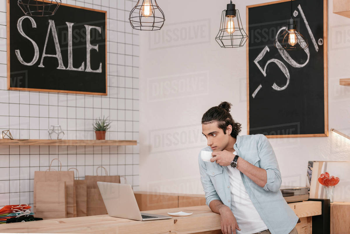 Handsome young shop owner using laptop and drinking coffee while ...
