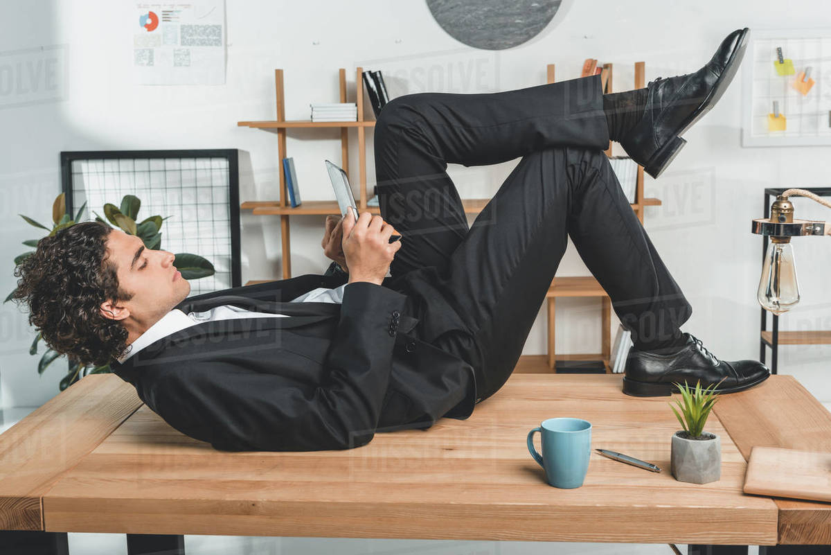 Side view of young businessman lying on table and using tablet in ...