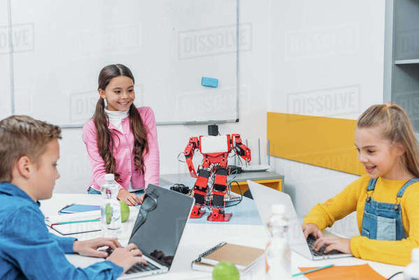 Smiling schoolchildren programming robot together and using laptops ...