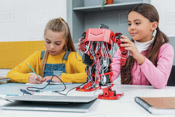 Schoolgirls writing at notebook and touching red electric robot during ...