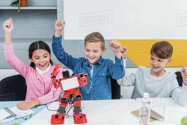 Happy schoolchildren raising hands and looking at red handmade robot at ...