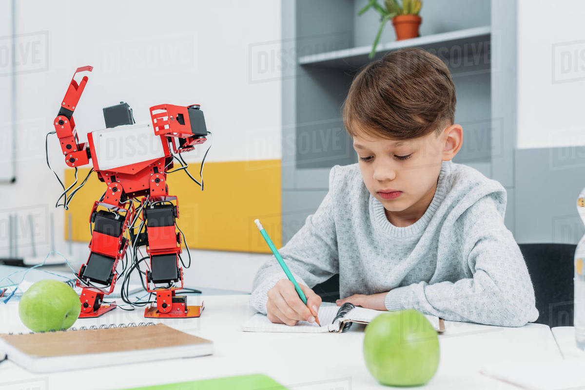 Focused schoolboy sitting at desk with robot model and writing in ...