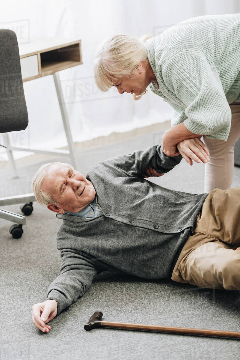 Old woman helping to stand up husband who fell down on floor with ...