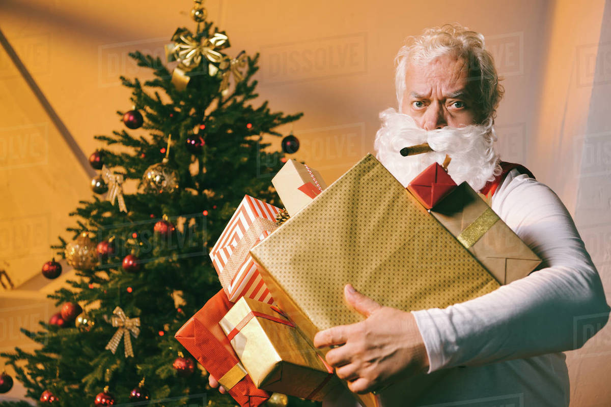 portrait of bad santa with cigar holding pile of gifts in room on ...