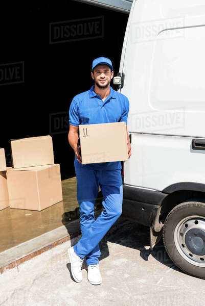 Smiling handsome delivery man with cardboard box leaning on van ...