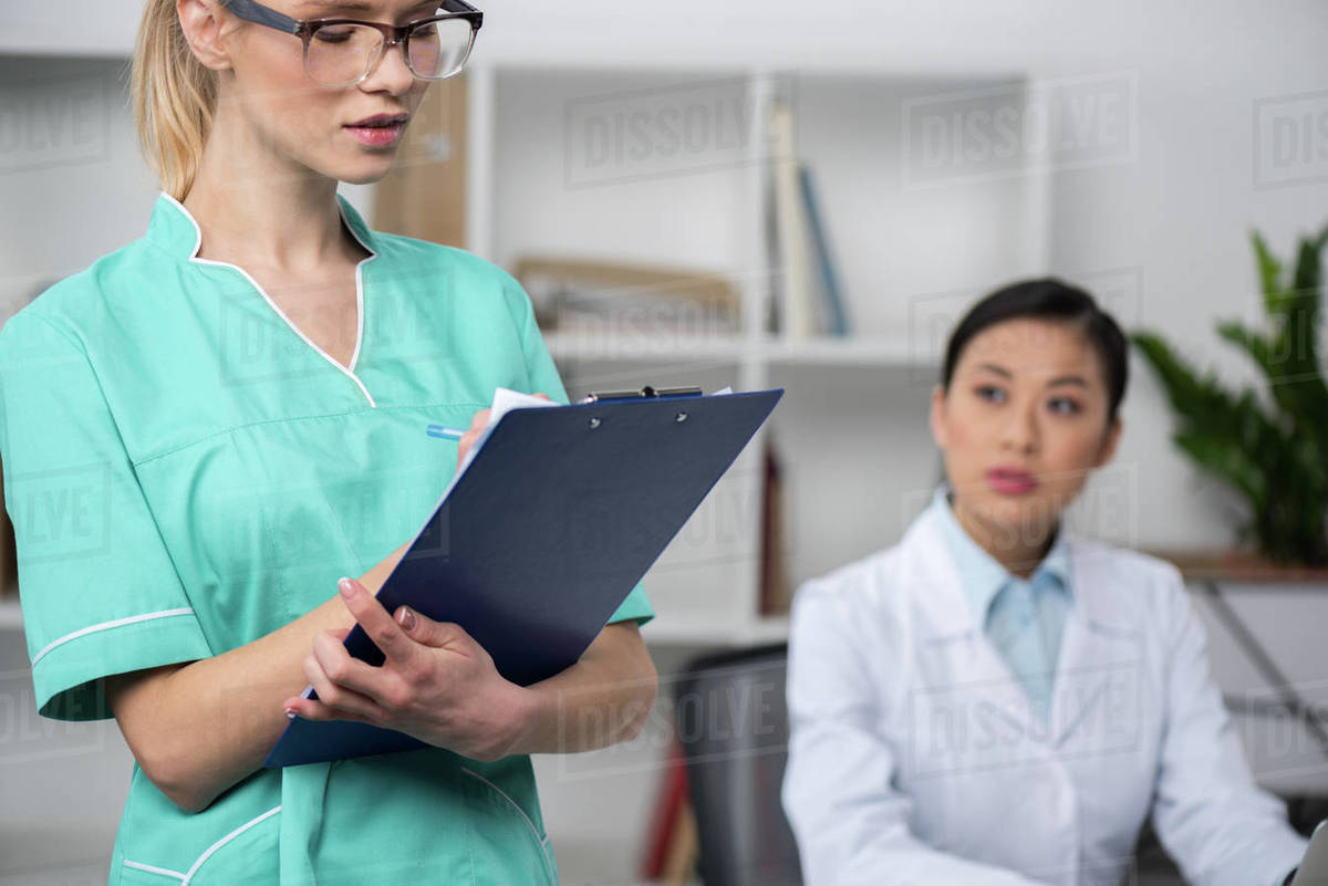 Portrait of focused doctor writing on clipboard with colleague behind ...