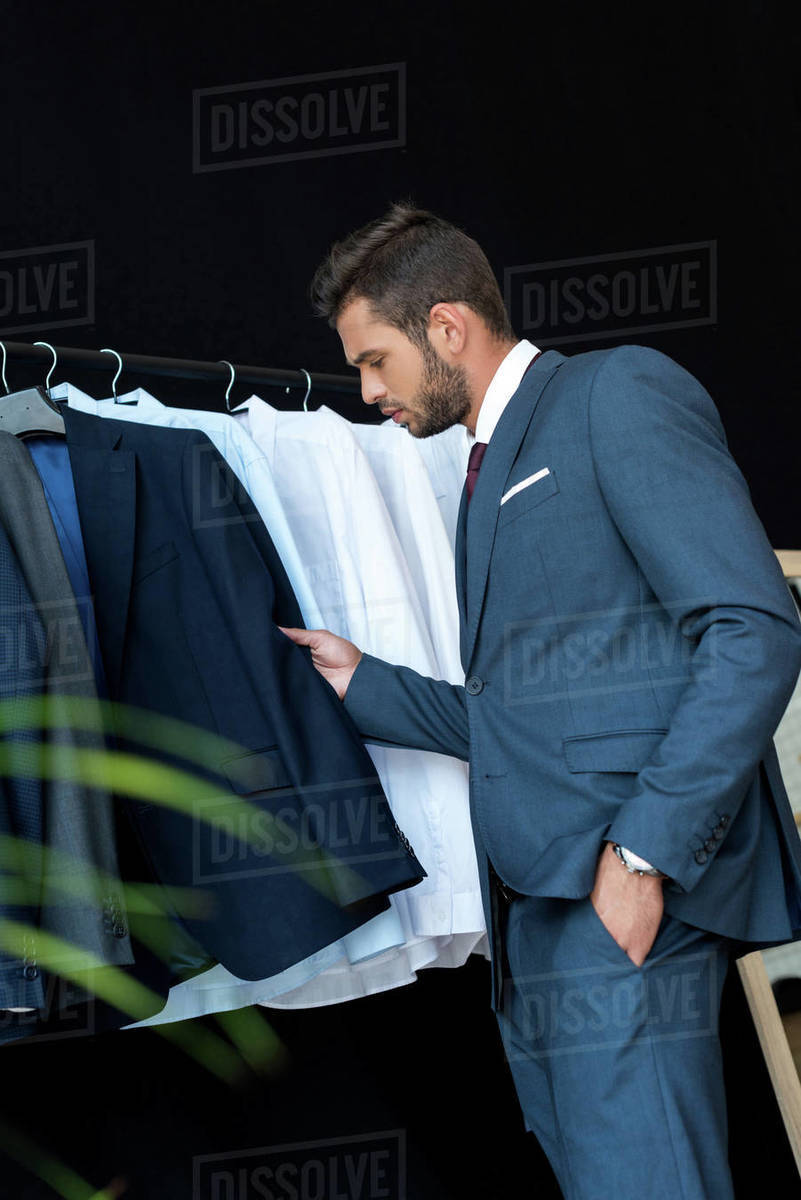Side view of handsome young businessman choosing suit in boutique ...