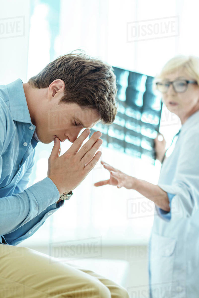 Disappointed young man praying as the doctor is revealing his diagnosis ...