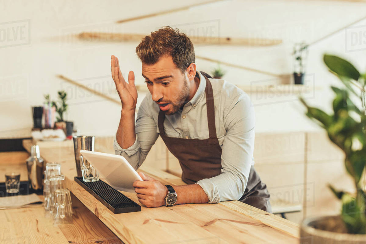 Portrait of shocked barista in apron using tablet while standing at ...