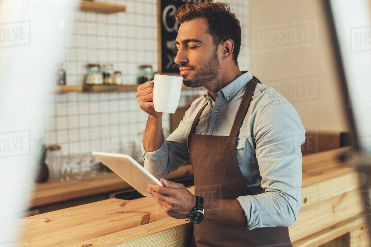 Side view of barista with cup of aromatic coffee using tablet in cafe ...