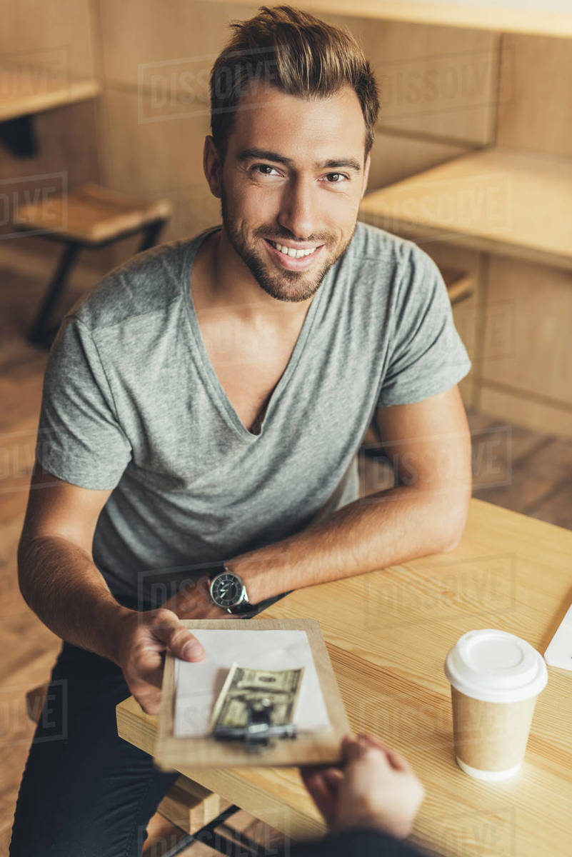 Portrait of smiling young man paying for order with cash in cafe ...
