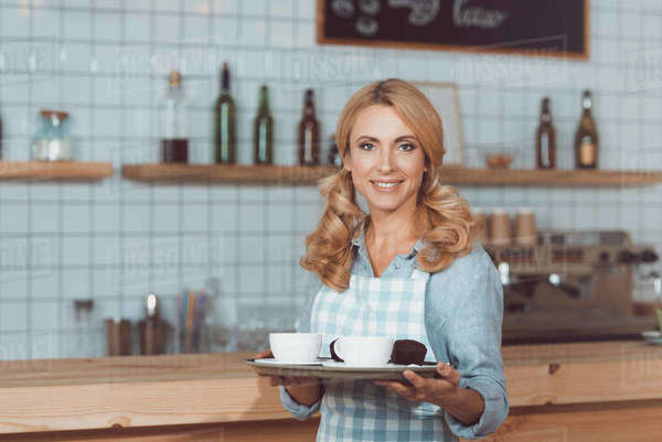 beautiful smiling waitress in apron holding tray with dessert and cups ...