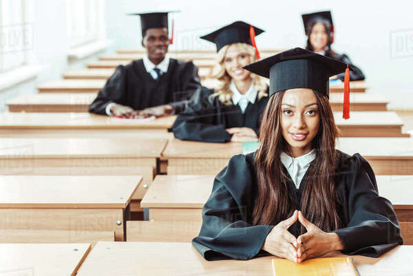 group of multiethnic students in graduation costumes sitting at class ...
