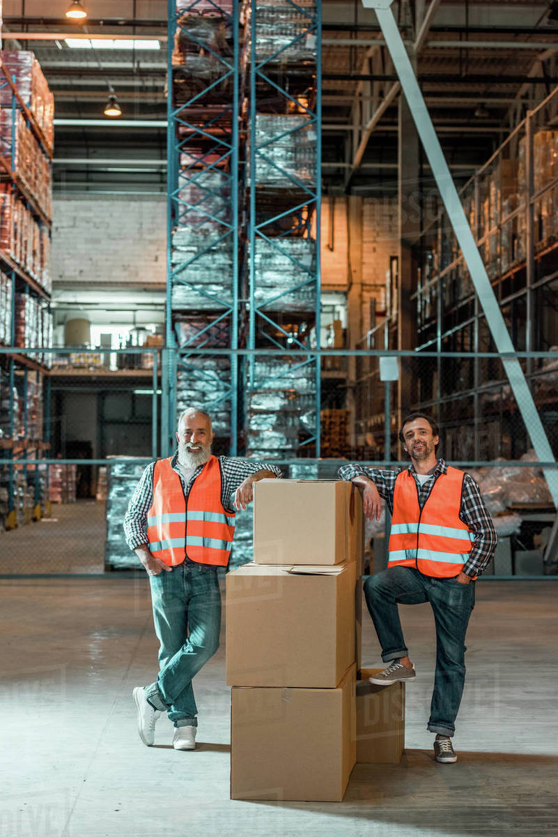 Male warehouse workers in vests standing with boxes and smiling at ...